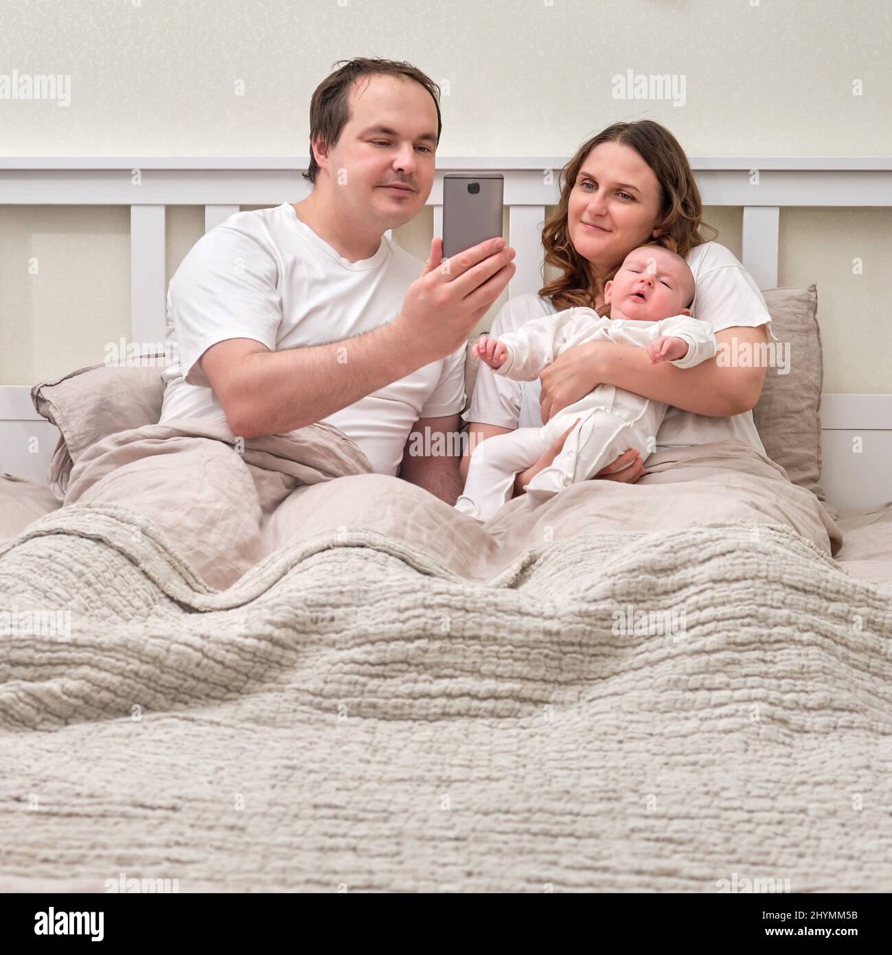 A man and a woman with a newborn baby on the bed take a selfie photo in ...