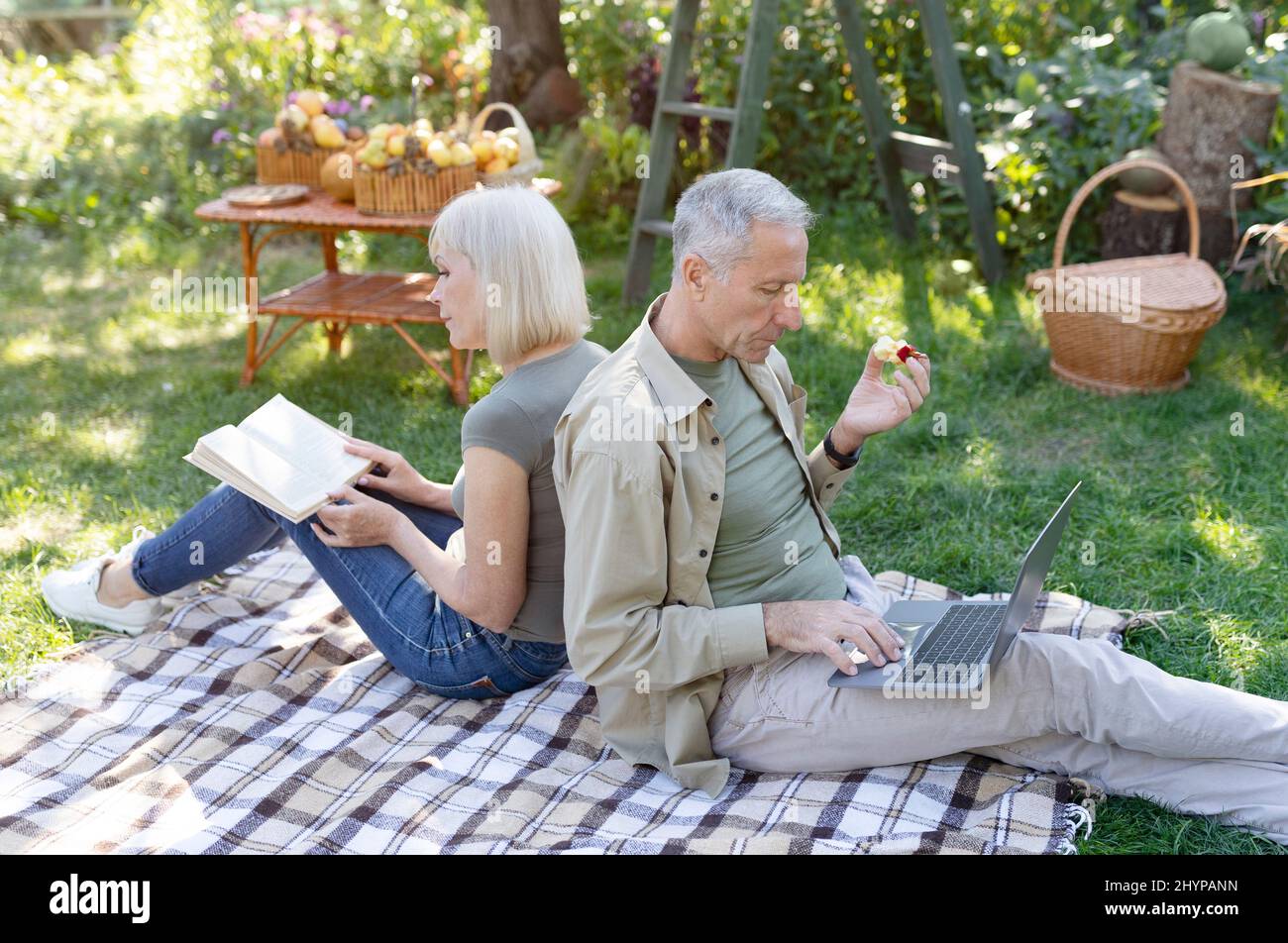 Modern elderly spouses resting in garden, man using laptop computer ...