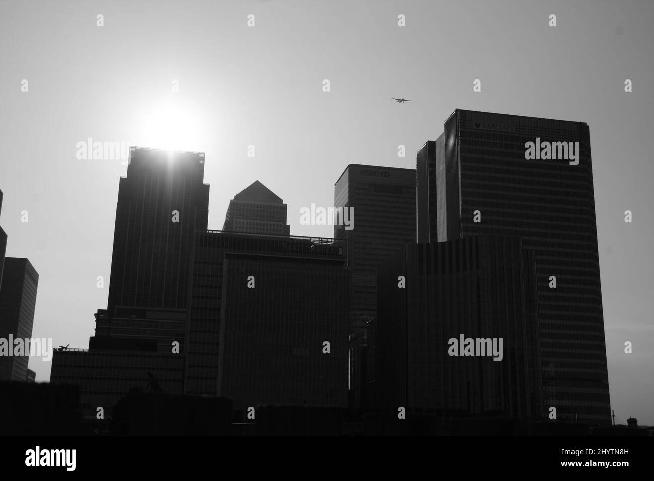 Grayscale low angle shot of the sun shining behind tall urban buildings ...
