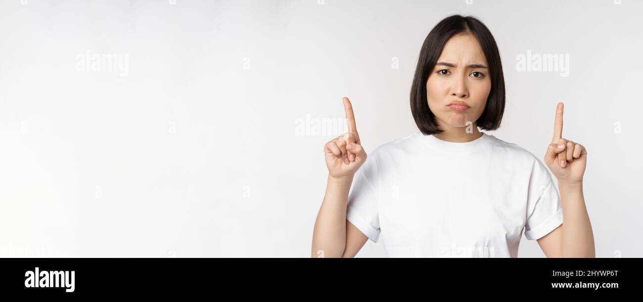 Sad and disappointed young asian woman pointing fingers up, showing ...