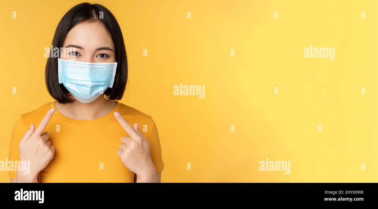 Portrait of smiling asian woman in medical face mask, pointing at her ...