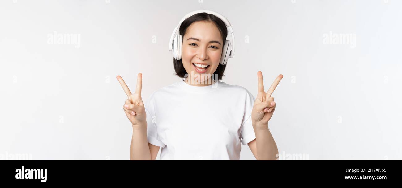 Happy asian woman wearing headphones and smiling, showing peace v-sign ...