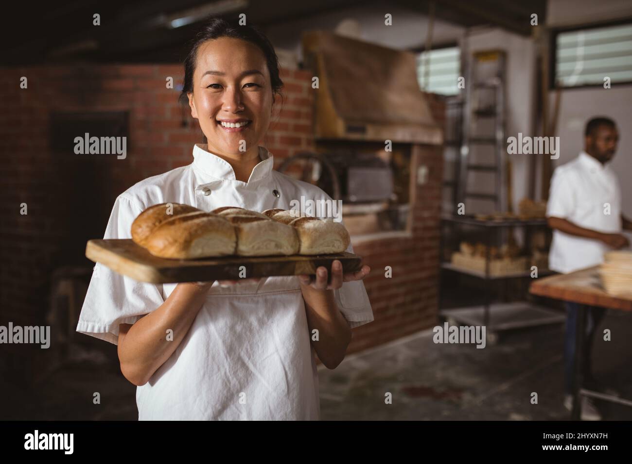 Portrait of smiling asian mid adult female baker holding serving board ...
