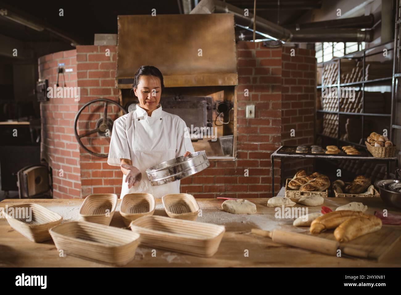 Mid adult asian female baker straining flour on table while working in ...