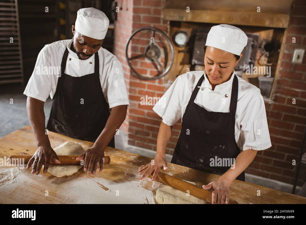 Multiracial mid adult male and female bakers rolling dough at table ...