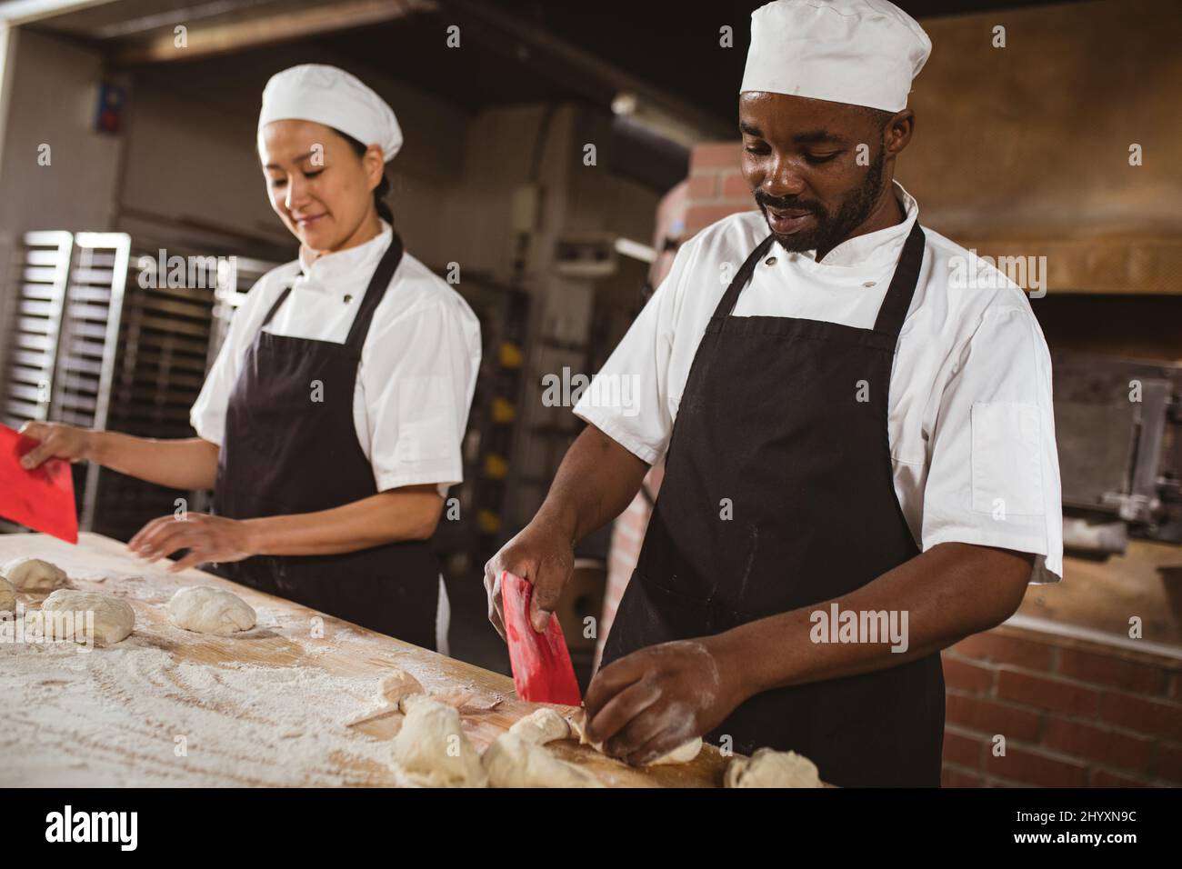 Multiracial mid adult male and female bakers cutting dough with dough ...