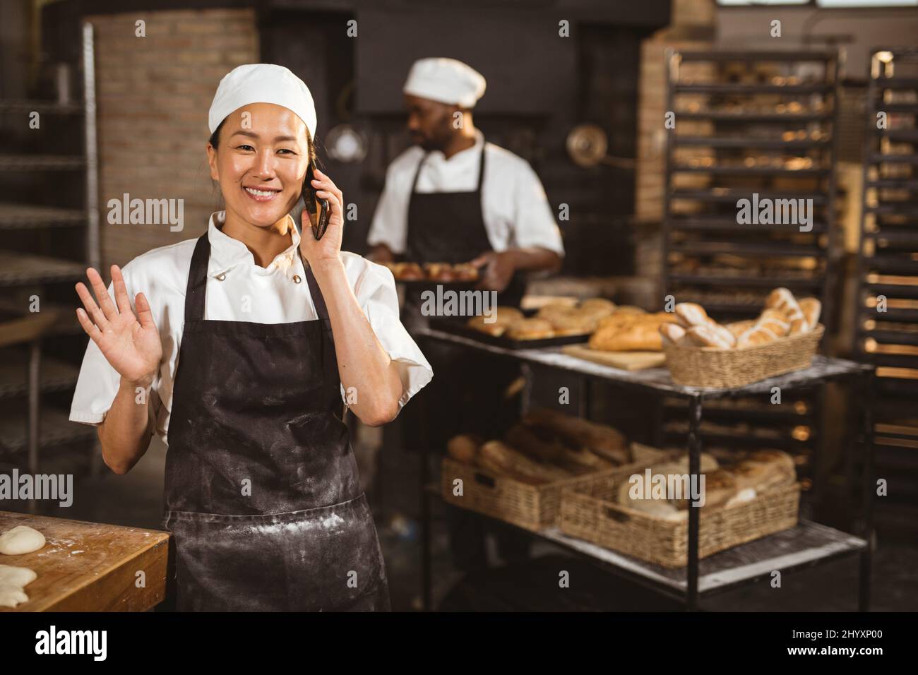 Smiling asian mid adult female baker talking on smart phone while ...