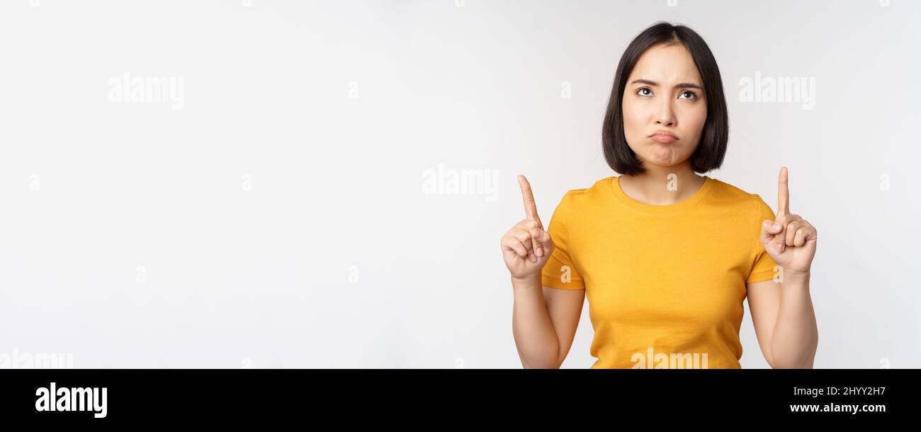 Disappointed asian woman looking, pointing fingers up with angry moody ...