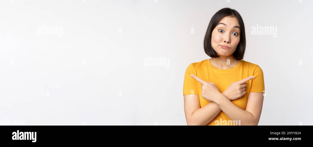 Complicated asian girl pointing fingers sideways, showing left and right choices, staring ...