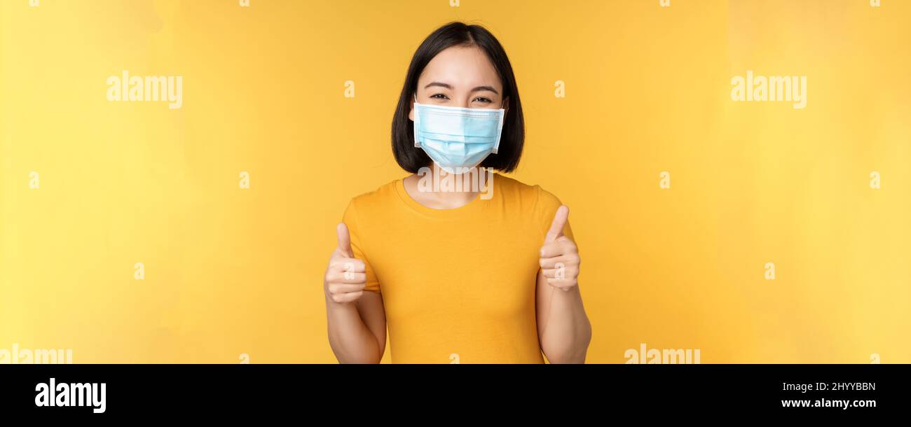 Smiling asian woman in medical face mask, showing thumbs up approval ...