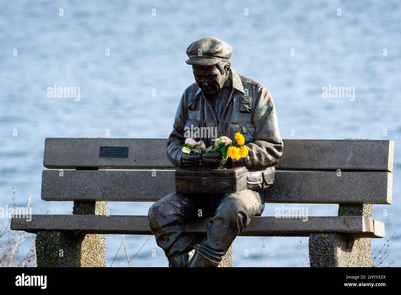Rusty statue of a man sitting on a bench holding flowers against a sea ...