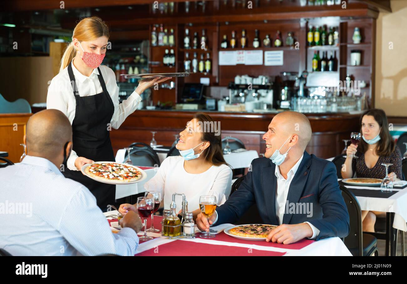Waitress in mask serving cheerful people in pizzeria Stock Photo - Alamy
