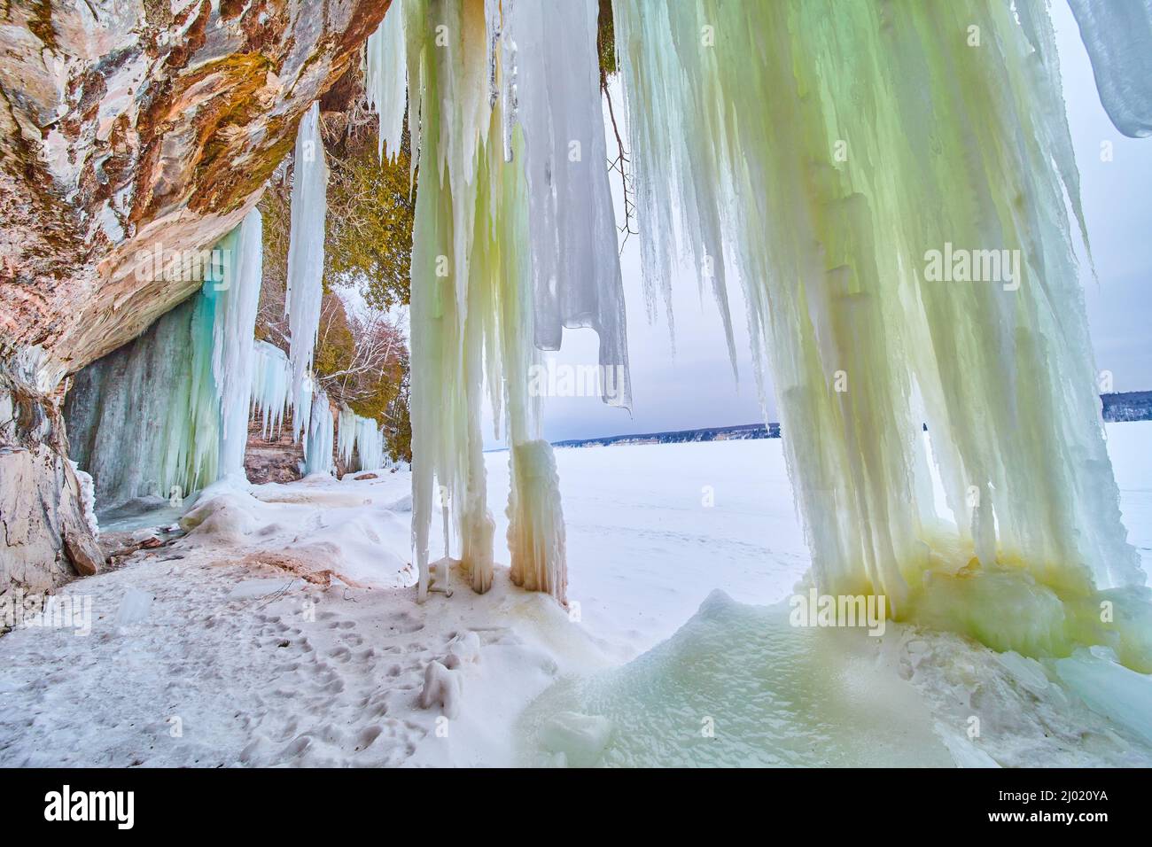 Thick icicle waterfalls of green and blue on rocks next to frozen lake ...