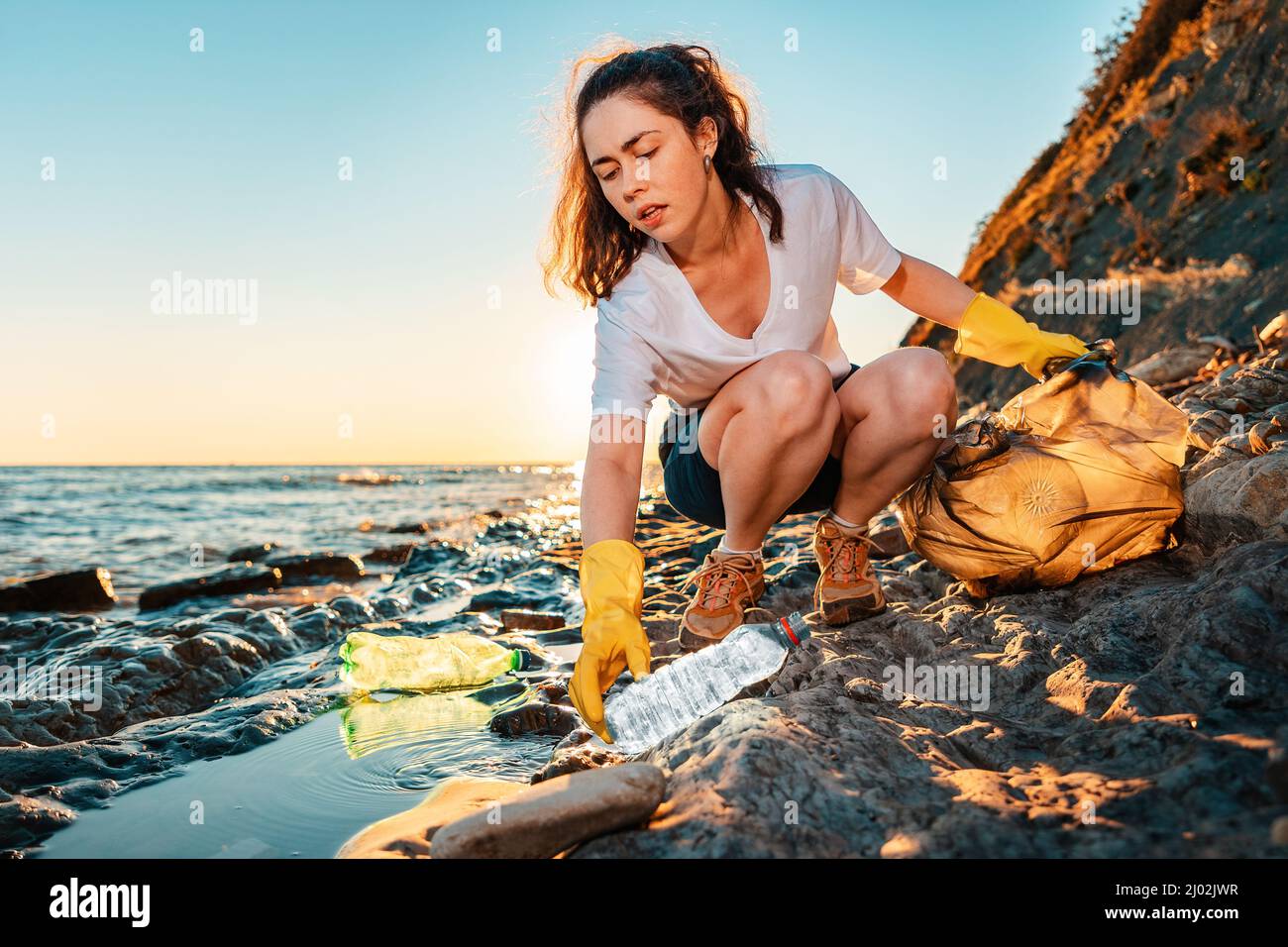 A young woman volunteer squats and picks up garbage on the ocean shore ...