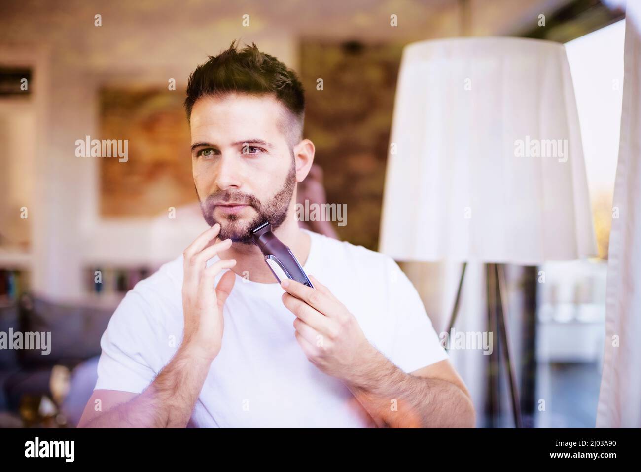 Portrait of a young man shaving his face with an electric razor at home ...
