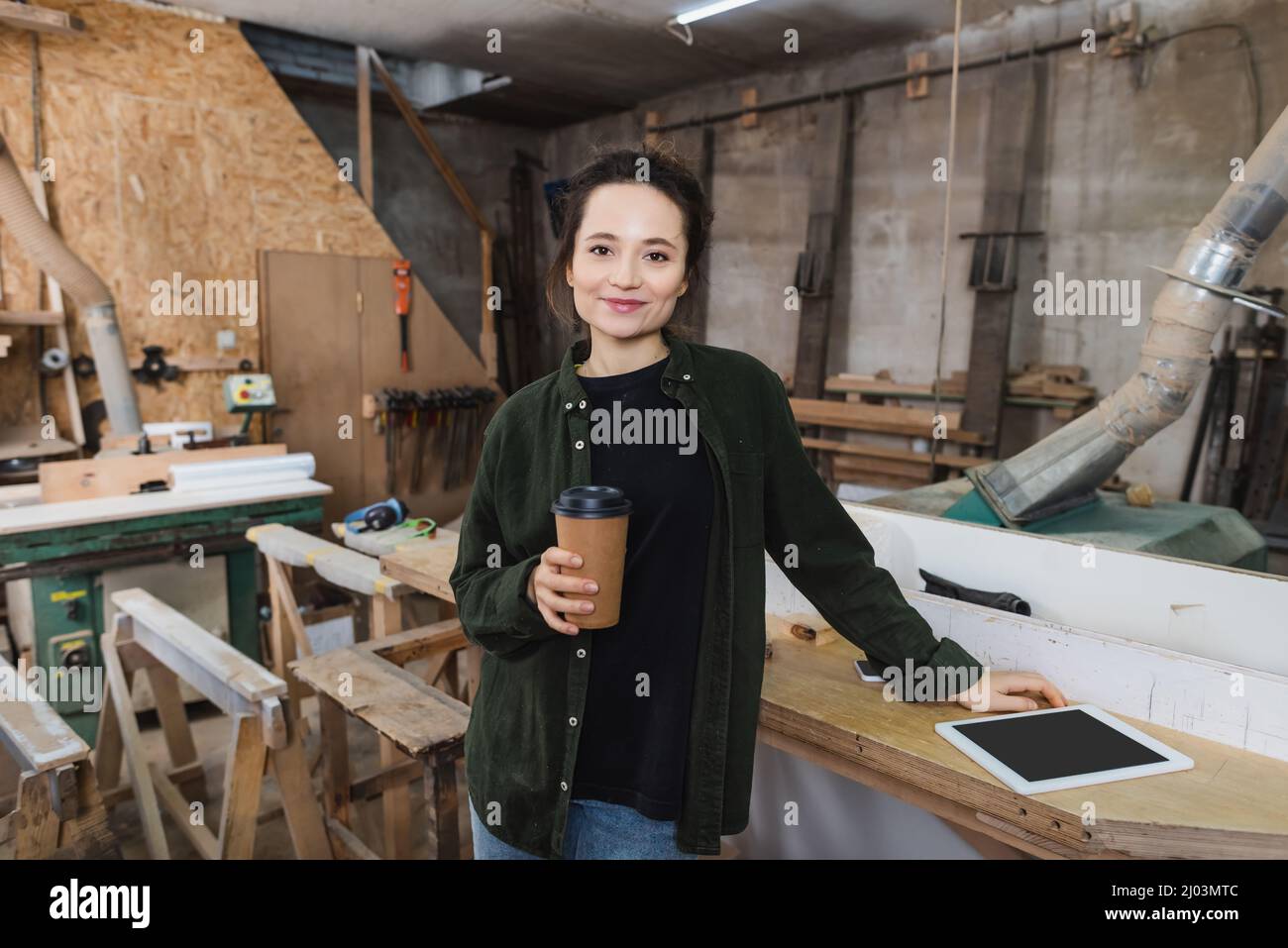 Smiling carpenter holding paper cup near devices in workshop Stock ...