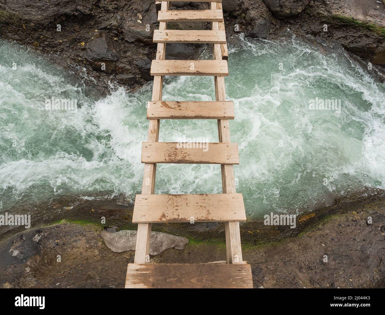A narrow wooden bridge over stormy icy river with rocky banks. View ...