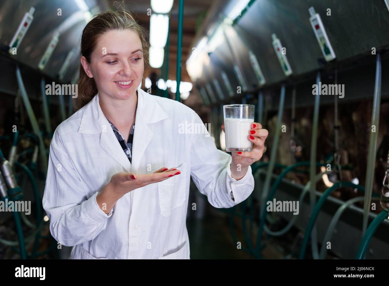 Female dairy engineer in white robe standing with glass of milk near ...
