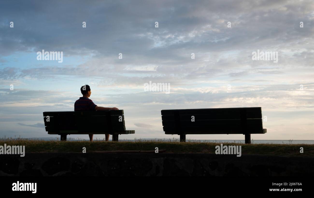 Man sitting on the bench facing the sea at dawn. Another empty bench by ...