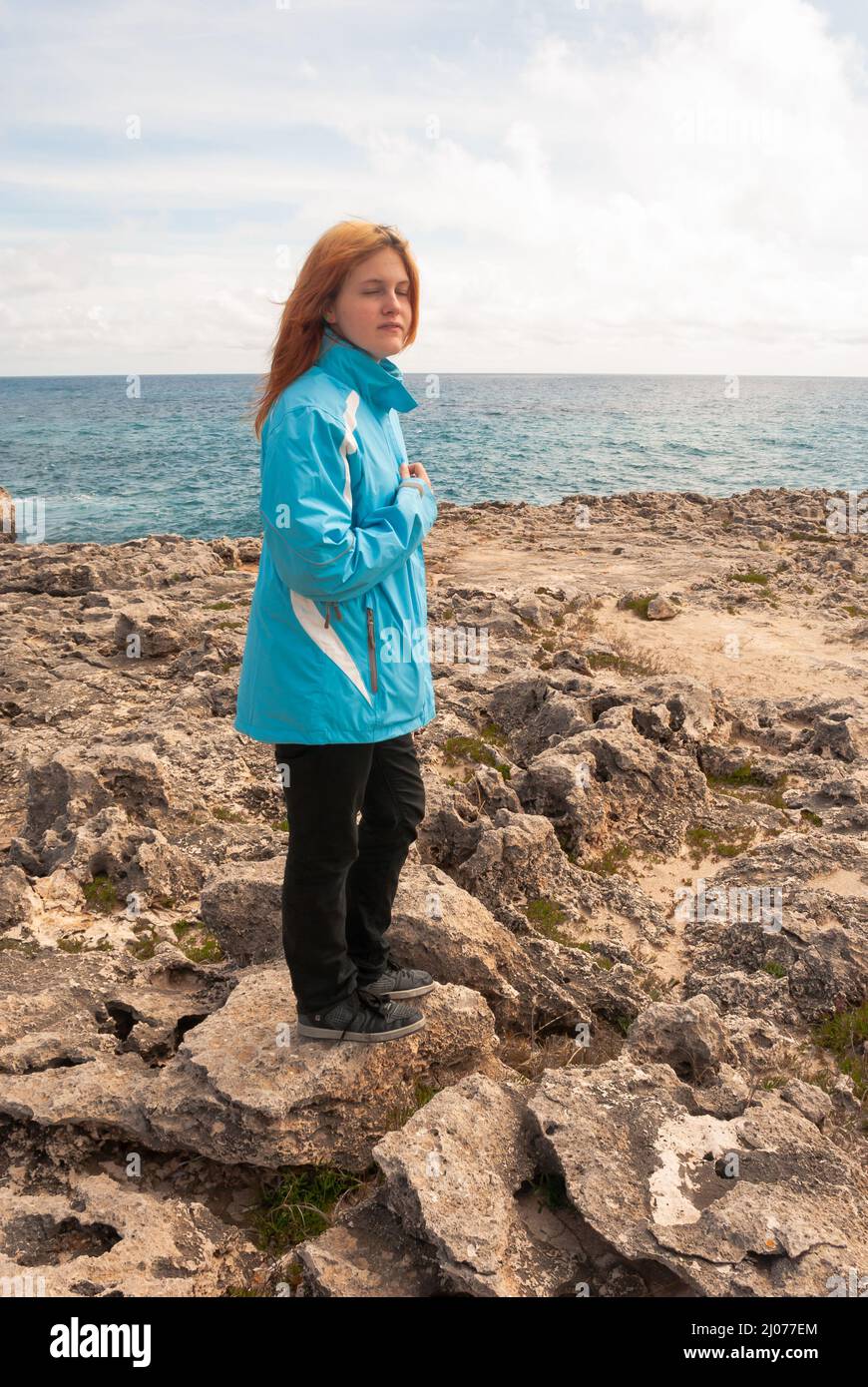 Young woman with orange hair, wearing a blue jacket, standing on a rock ...