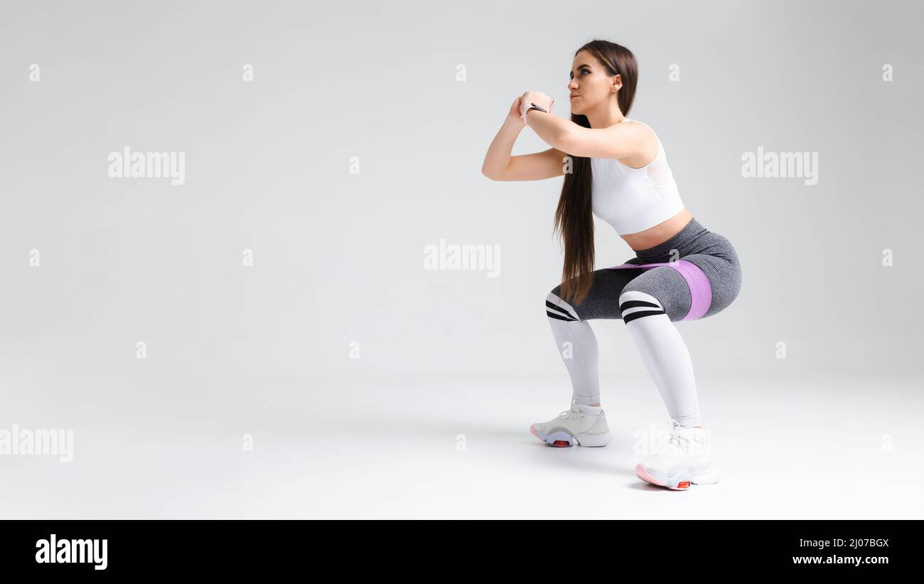 Athletic woman in sports wear squatting with resistance band at studio ...