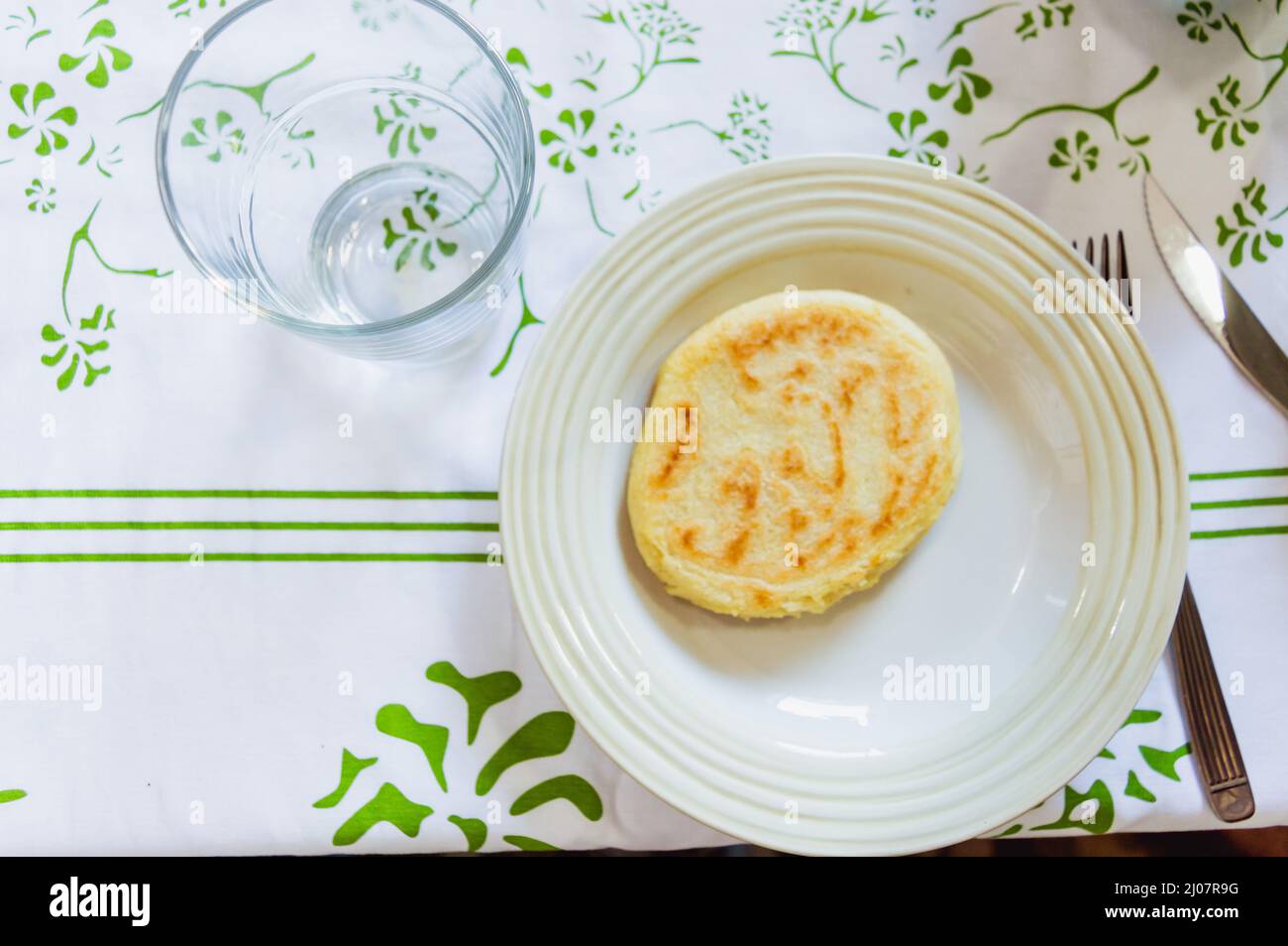 single arepa in a porcelain plate on the table next to a glass cup ...