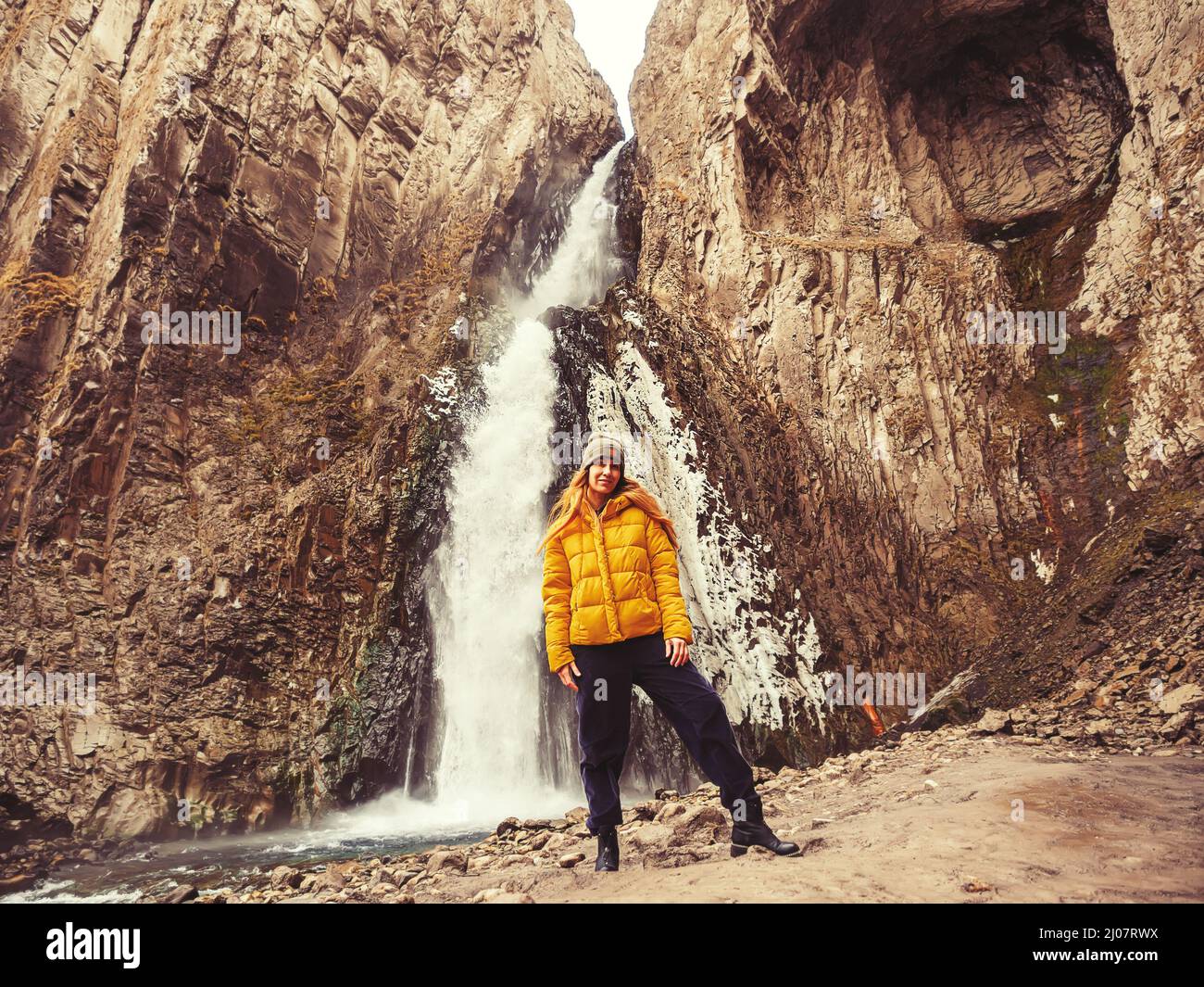 A smiling woman stands against the background of icy stormy waterfall ...