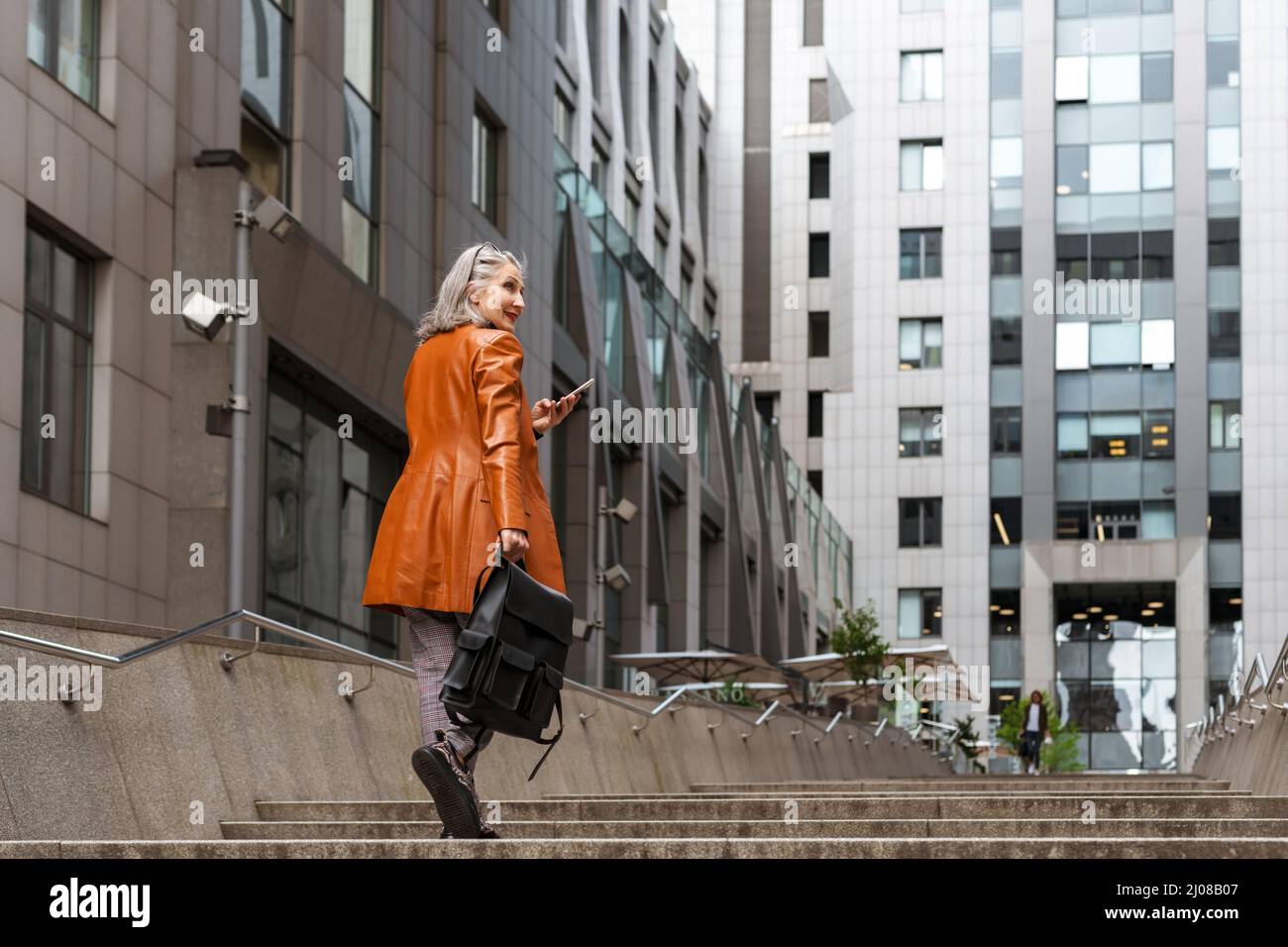 Grey senior woman using mobile phone while going up stairs outdoors ...