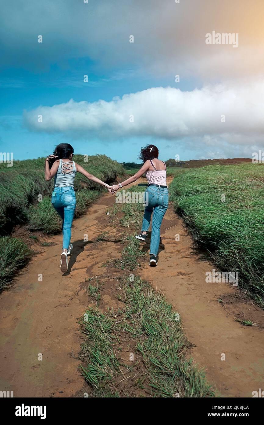 Two Latina teen friends run together holding hands down a country road ...