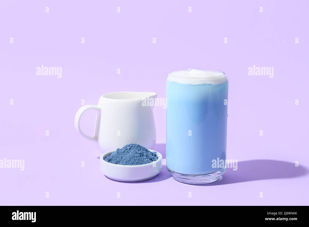 Glass of blue matcha tea, jug with milk and bowl with powder on purple ...