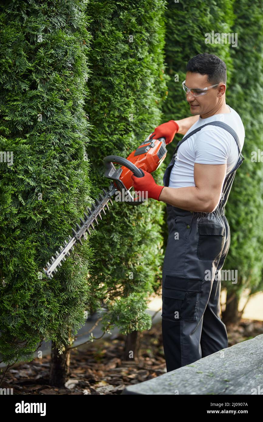 Strong male gardener in uniform, glasses and gloves using electric ...
