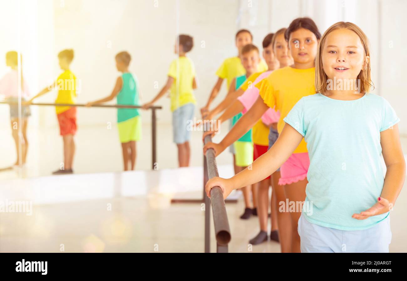 Smiling tween girl practicing classic dance moves near ballet barre ...