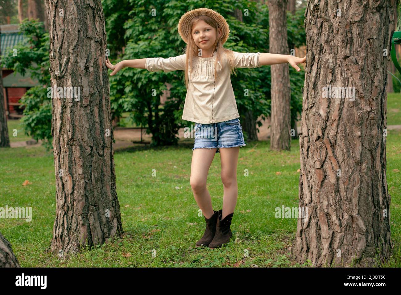 Cheerful tween girl posing between two tall trees in green summer park ...