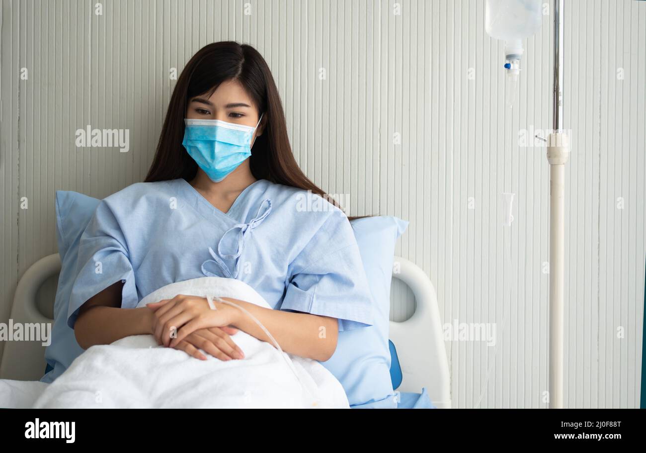 Sad Asian patient woman lying on the hospital bed and wearing a face mask to protect coronavirus ...