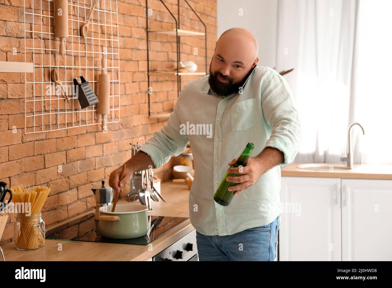 Bald man drinking beer and talking by phone while cooking in kitchen ...