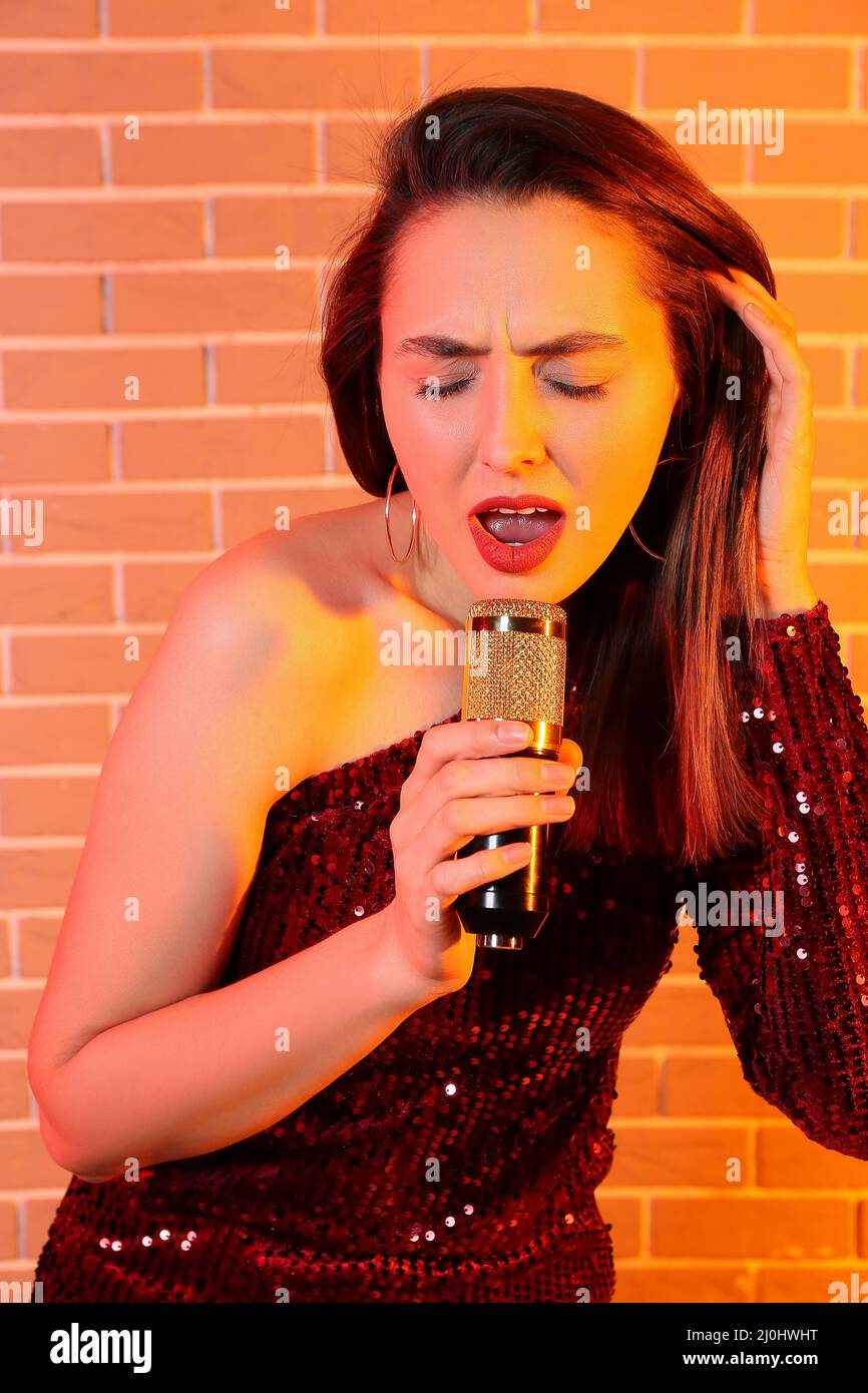 Pretty young singing woman with microphone on color brick background ...