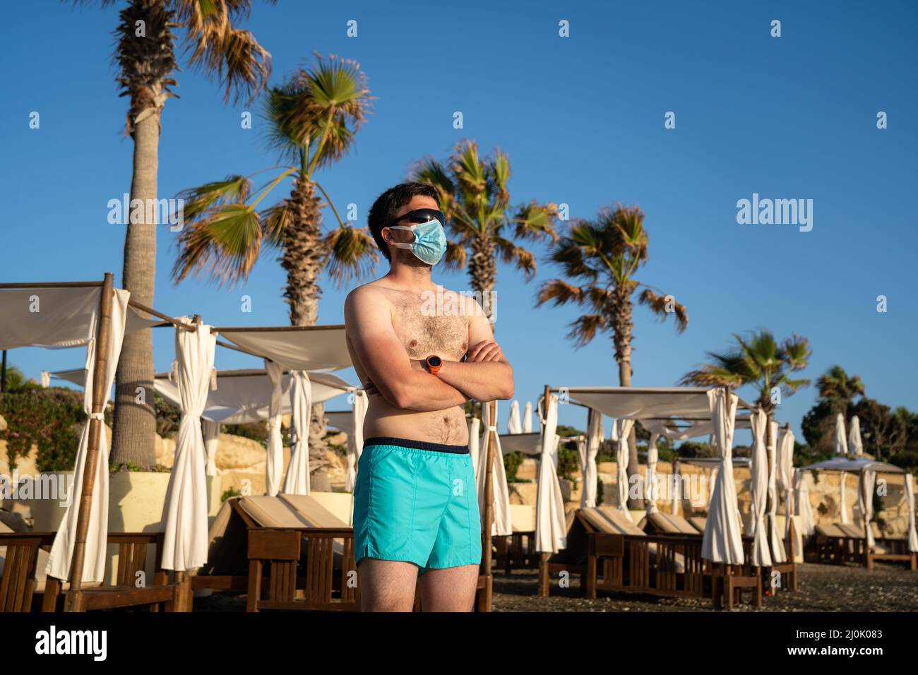 Man in medical mask stands alone on empty beach near sun loungers and ...