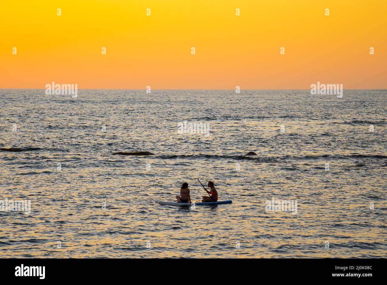 Two women sharing paddle board at sunset on calm waters in ...