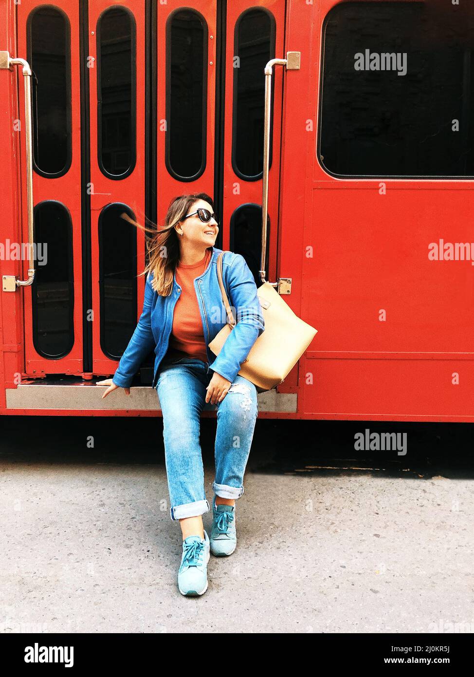 Happy young woman traveler in denim clothes sitting on steps of red ...