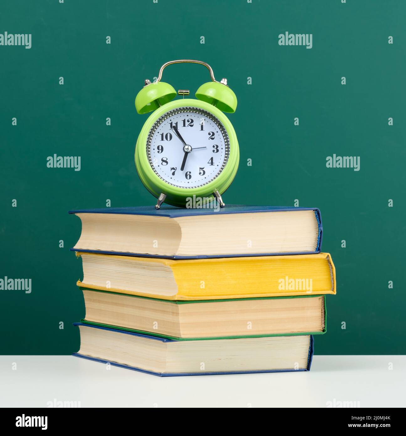 Round alarm clock and stack of books on empty chalk green board ...