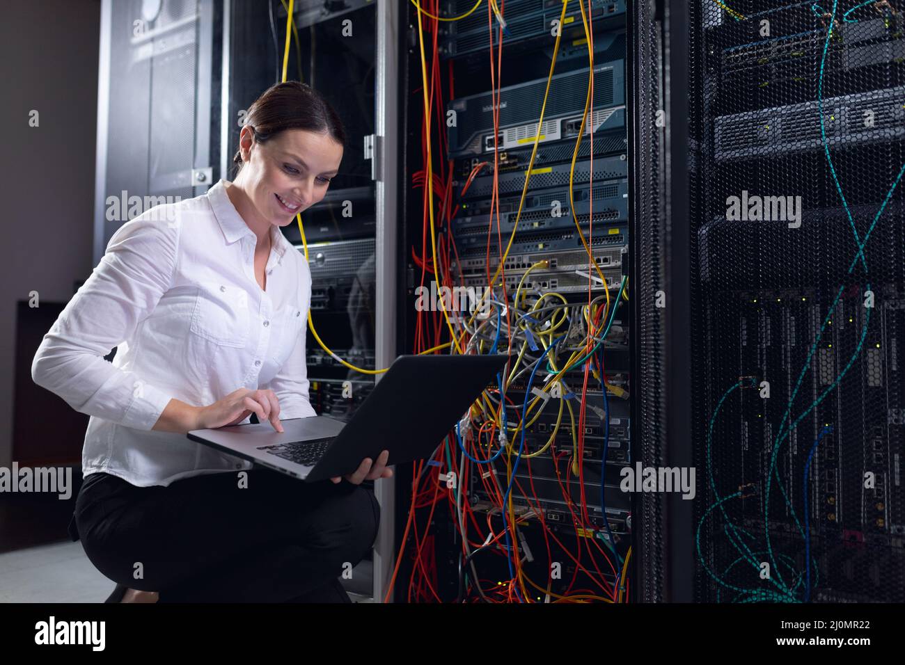 Caucasian female engineer using laptop and inspecting computer server in computer server room ...