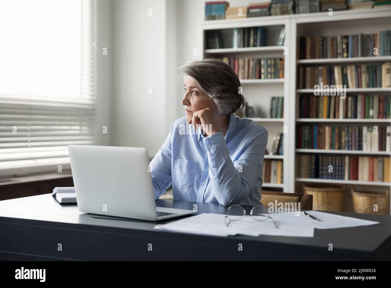 Serious older businesswoman sit at workplace desk staring out window ...