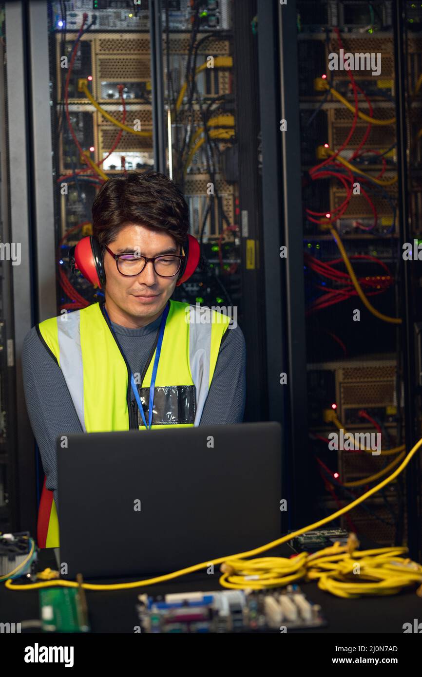 Asian male engineer wearing ear plugs using a laptop in computer server ...
