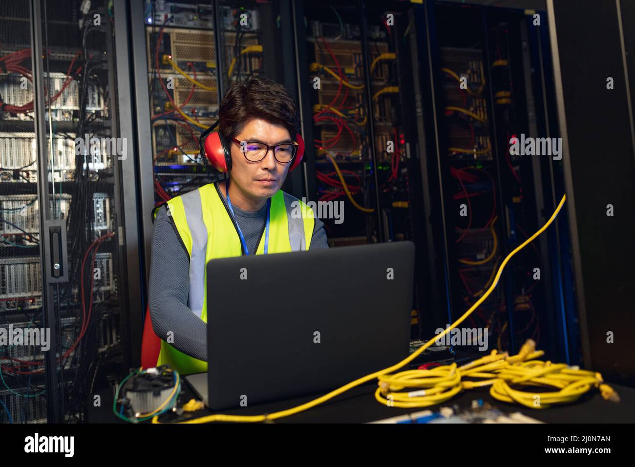 Asian male engineer wearing ear plugs using a laptop in computer server ...