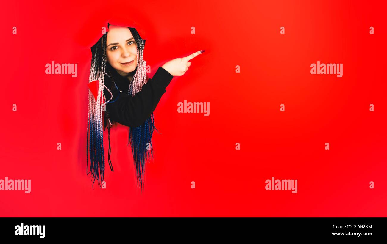 Young woman sticking out of hole of red background and pointing index ...