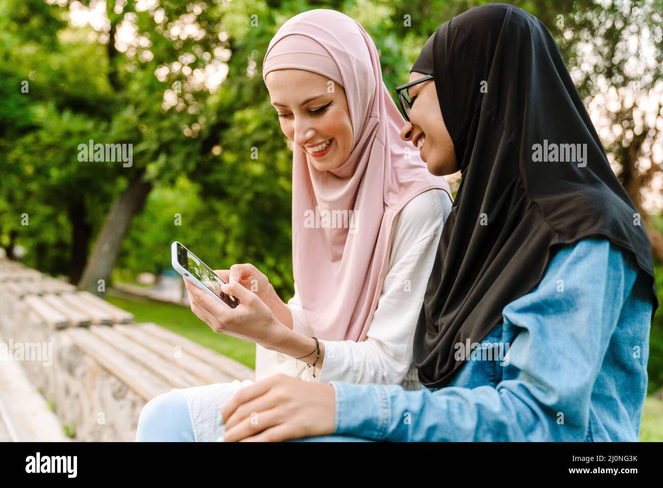 Two happy young muslim women in hijab sitting on a bench looking at ...