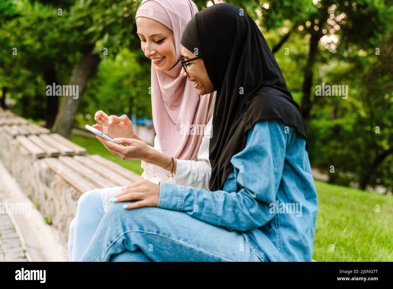 Two happy young muslim women in hijab sitting on a bench looking at ...