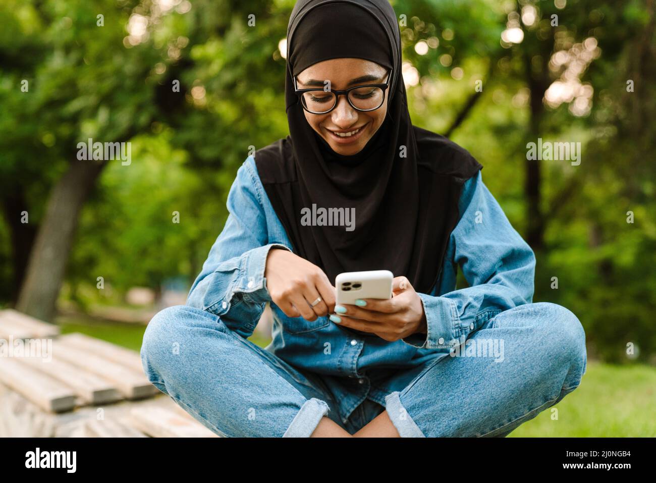 Happy young muslim woman in hijab sitting on a bench using mobile phone ...
