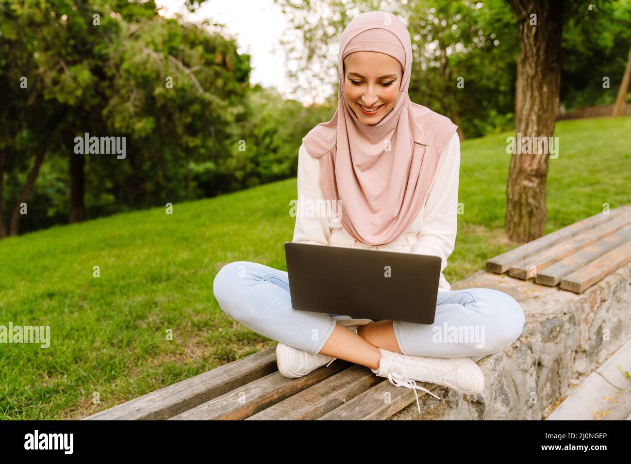 Happy young muslim woman in hijab sitting on a bench with laptop ...