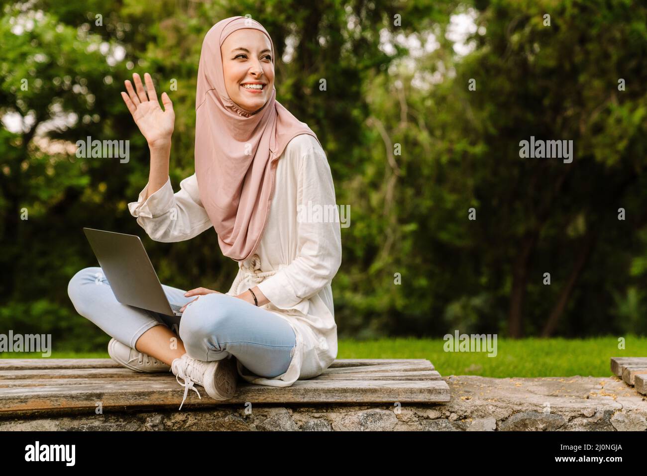 Happy young muslim woman in hijab sitting on a bench with laptop ...